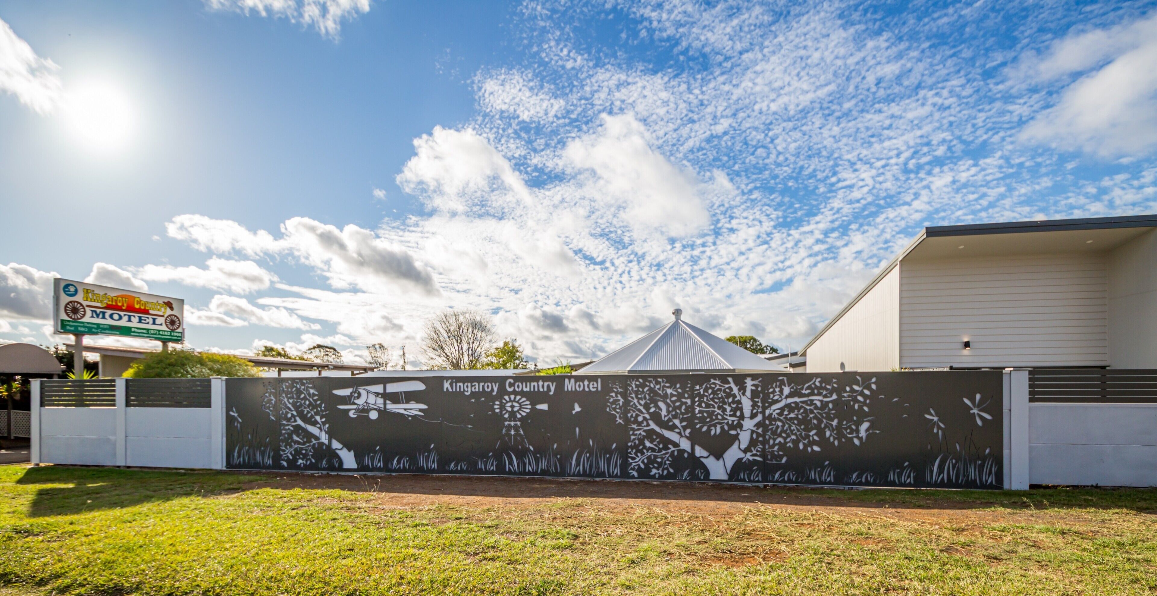 Um motel com um mural de uma árvore e um avião, uma placa indicando o motel, e um edifício com fachada branca.