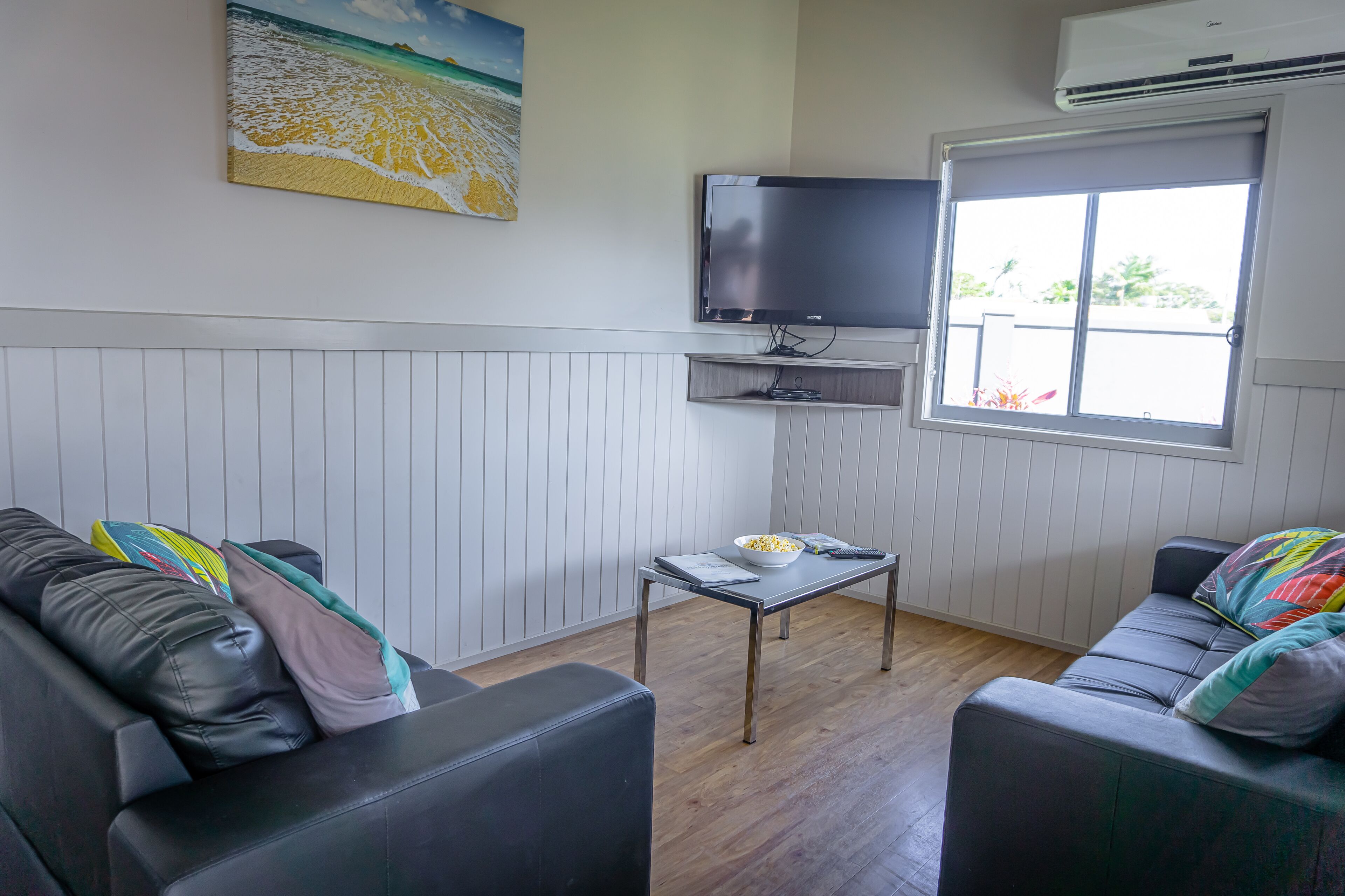 Uma sala de estar com sofá, mesa de centro, TV de tela plana e janela com vista para árvores.