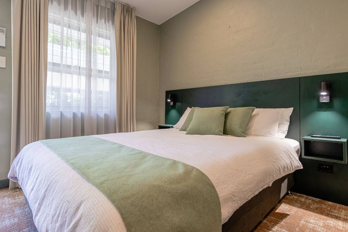 Uma cama bem arrumada com colcha verde e branca, cabeceira com detalhe verde e mesa de cabeceira com controle remoto.