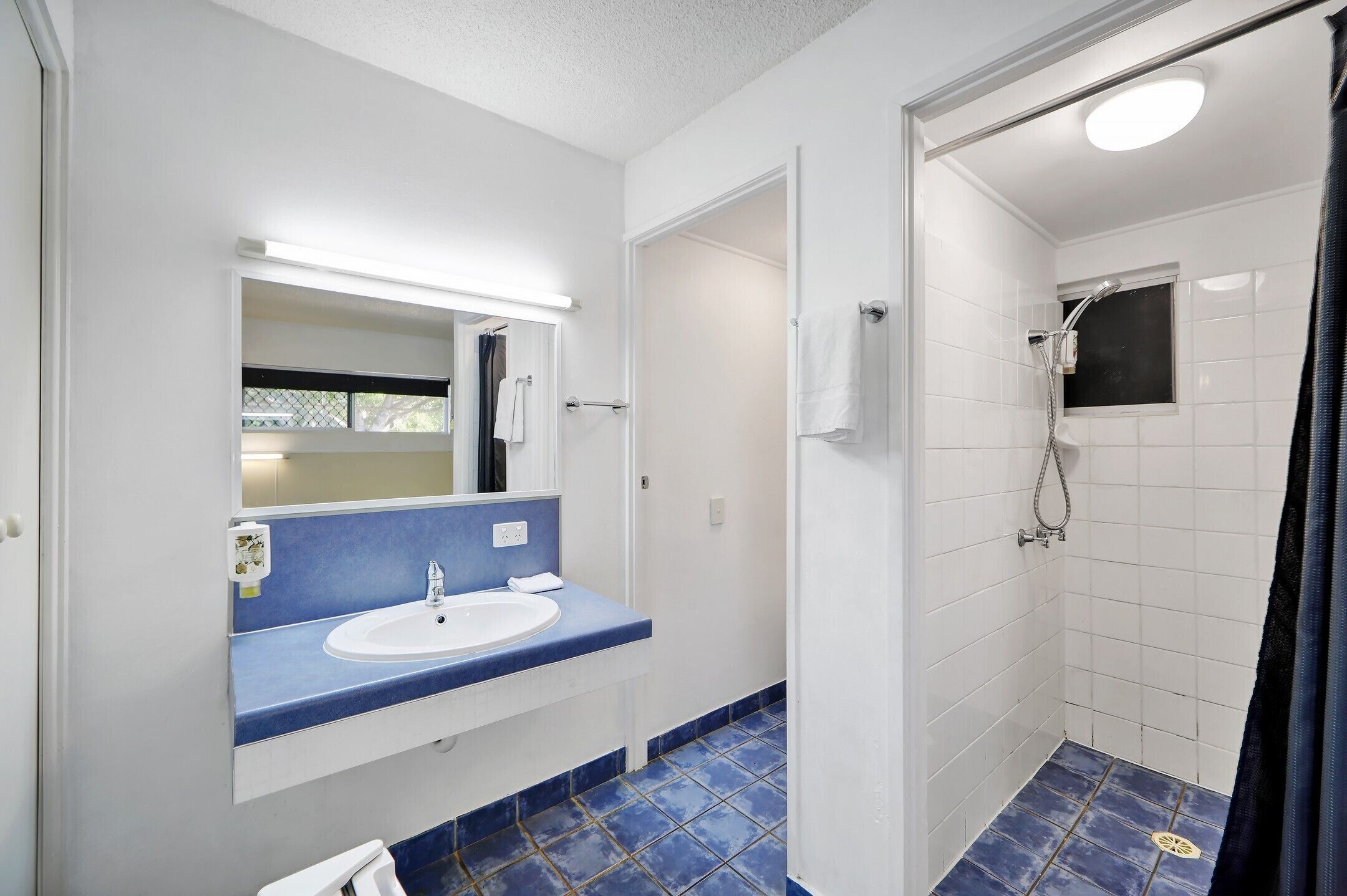 Casa de banho com lavatório branco, bancada azul e chuveiro com azulejos brancos.