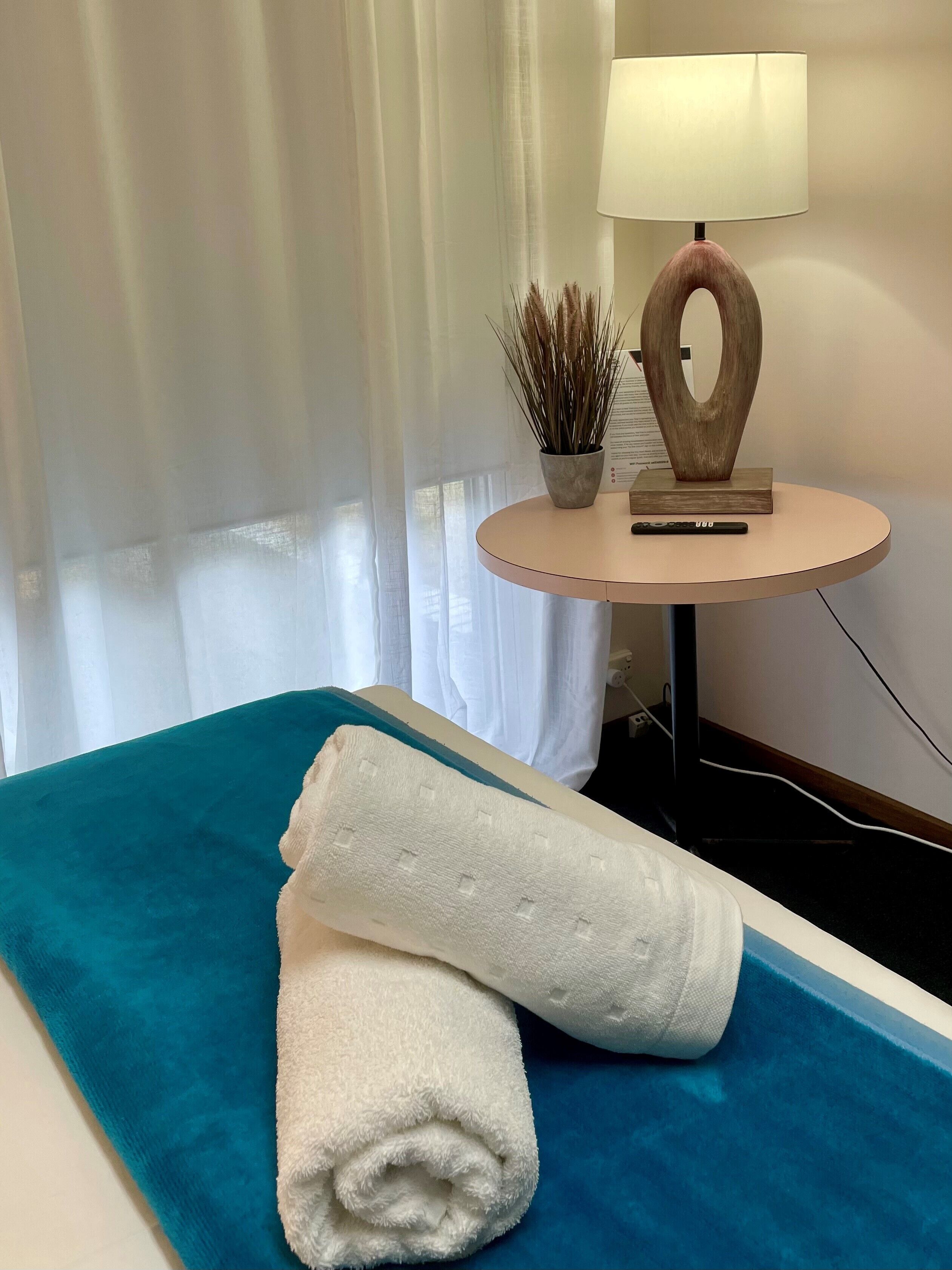 Cama bem arrumada com um edredom azul e duas toalhas brancas dobradas, uma mesinha com abajur e controle remoto, e um vaso de planta.