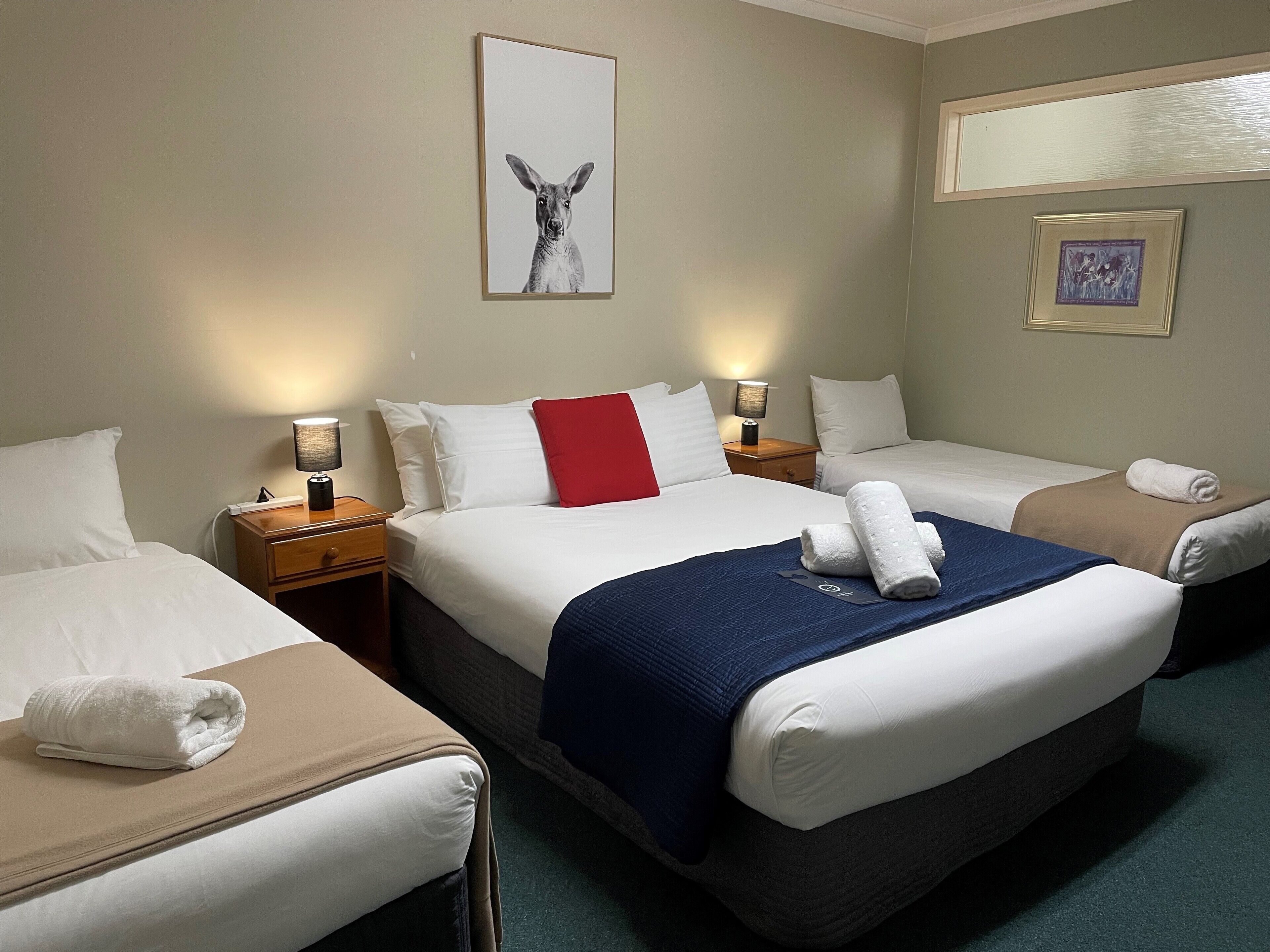 Quarto de hotel com duas camas de solteiro e uma cama de casal, cada uma com roupa de cama branca e uma almofada decorativa vermelha. Duas mesas de cabeceira com luminárias, um quadro na parede e uma toalha em cada cama.