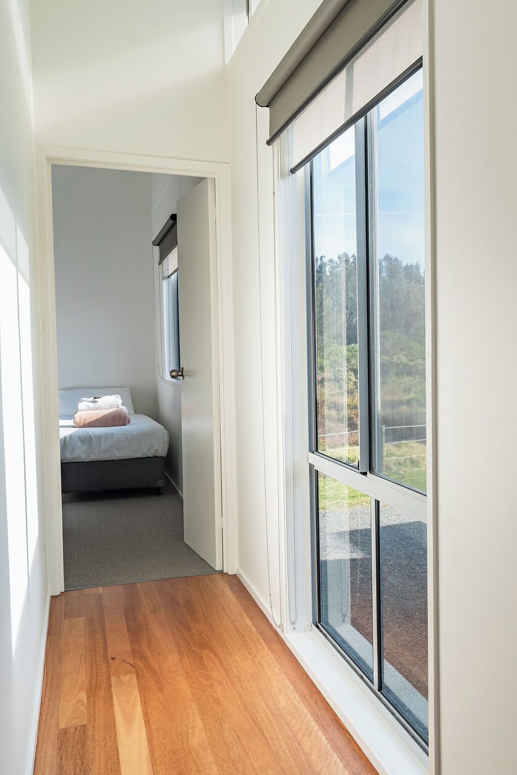 Um corredor estreito com uma janela grande, uma cama com roupa de cama branca e piso de madeira.