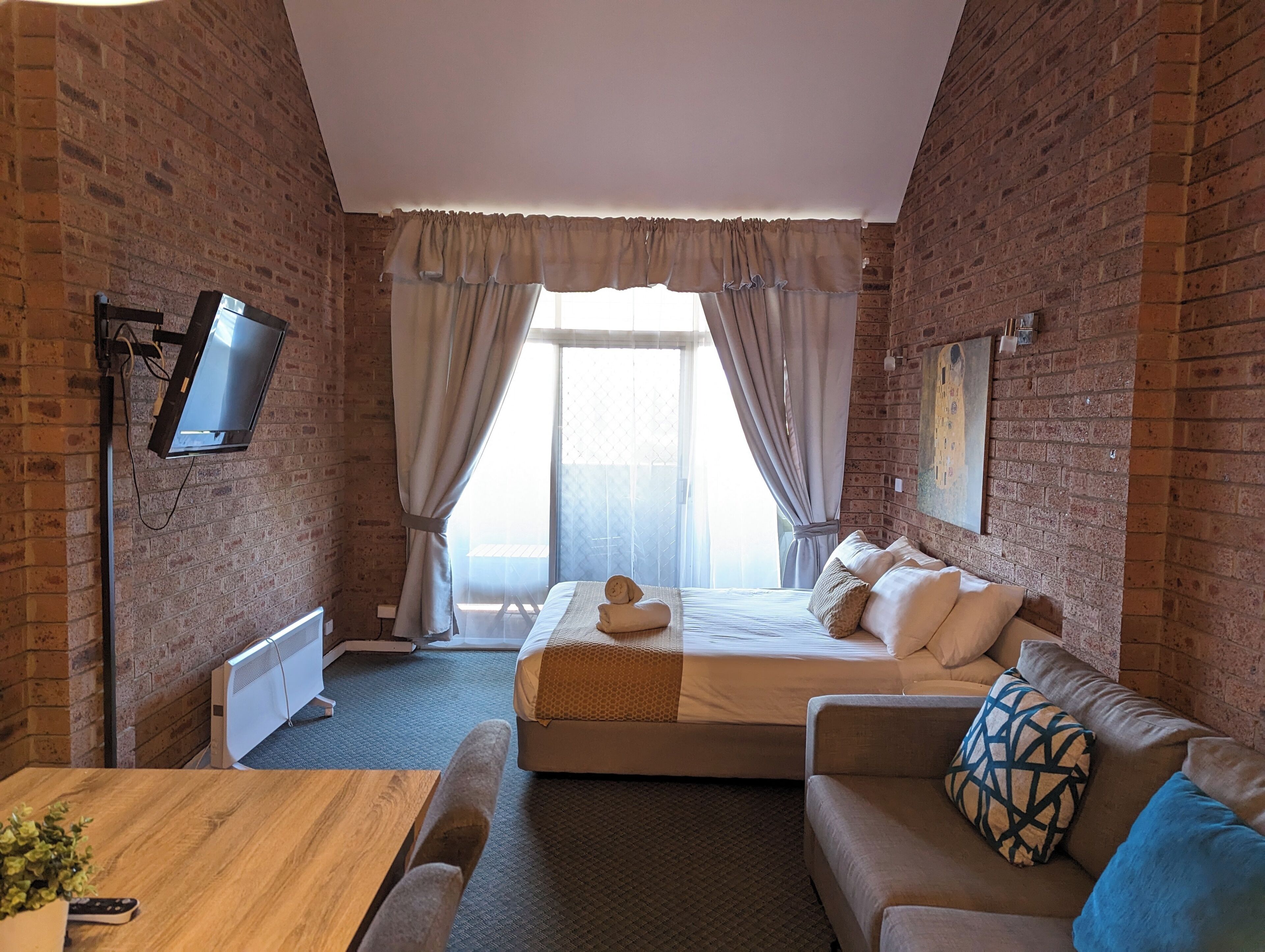 Quarto de hotel com parede de tijolos, televisão de tela plana na parede, cama com manta bege, sofá com almofada estampada e uma pequena mesa com uma planta.