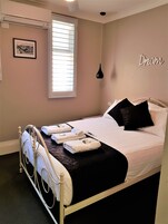 Uma cama bem arrumada com lençóis brancos e almofadas pretas, uma mesa de cabeceira com abajur e um quadro na parede.
