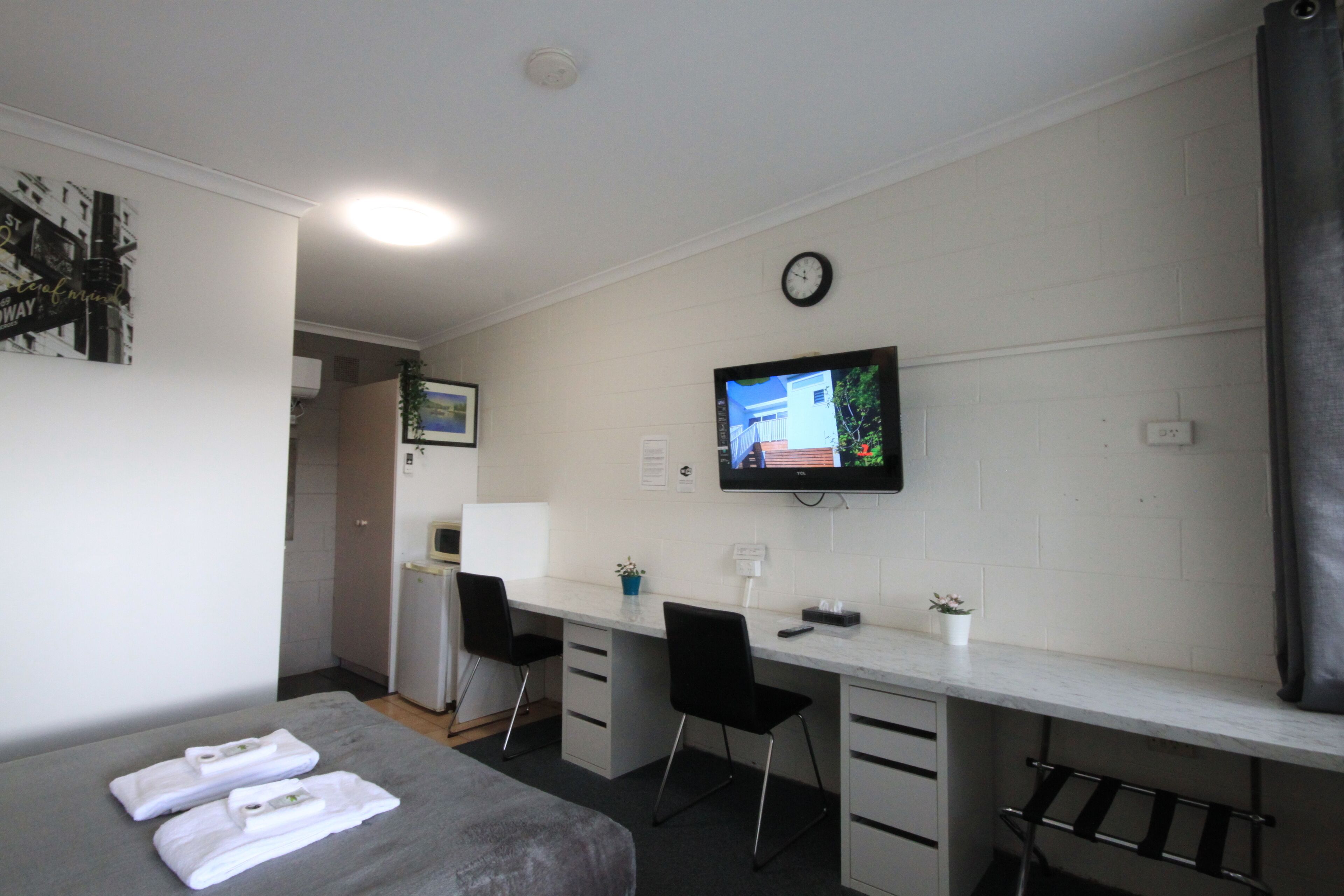 Quarto de hotel com cama, escrivaninha com duas cadeiras, televisão e pequena cozinha.