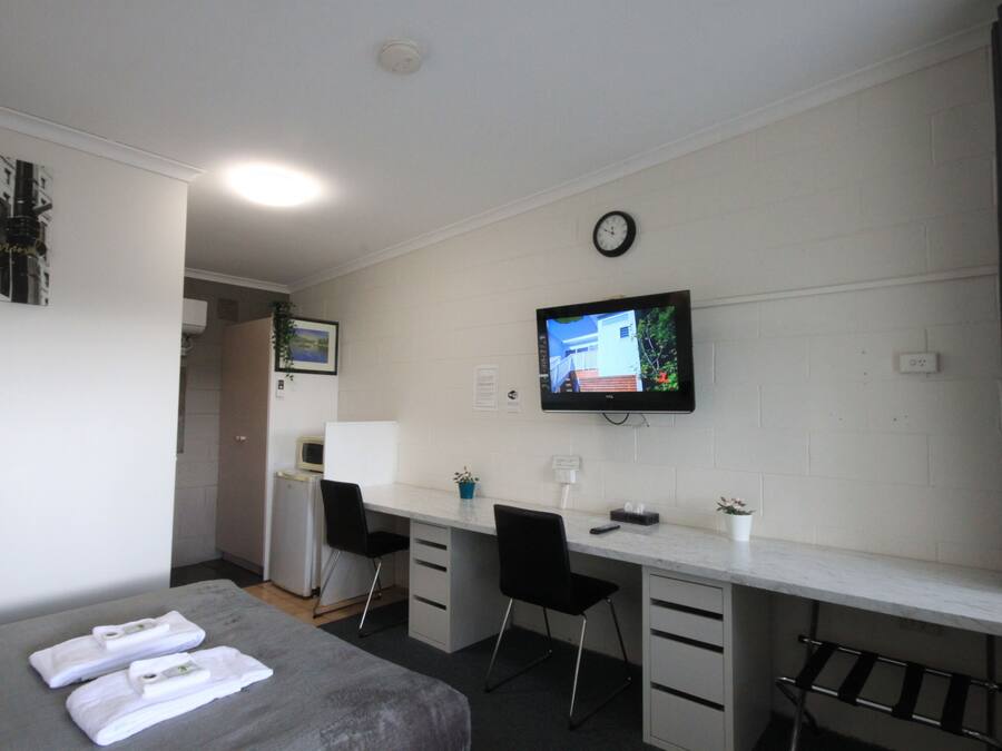 Quarto de hotel com cama, escrivaninha com duas cadeiras, televisão e pequena cozinha.