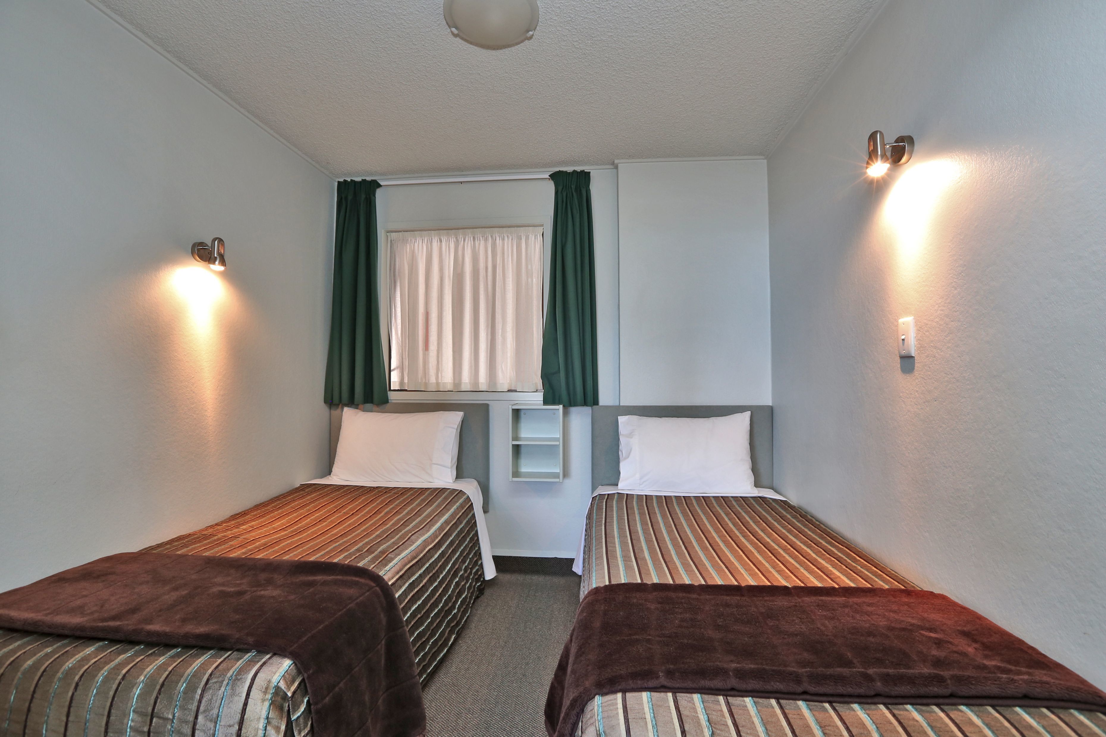 Quarto de hotel com duas camas de solteiro, uma janela com cortinas e luminárias fixadas na parede.