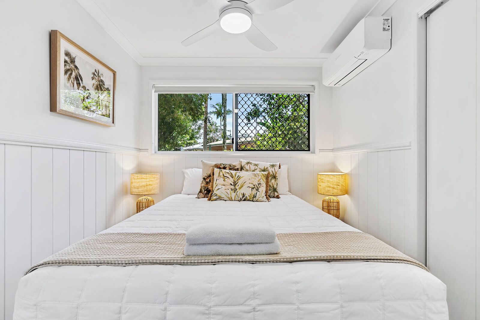 Quarto com cama, duas luminárias de cabeceira, ventilador de teto e janela com vista para o verde.