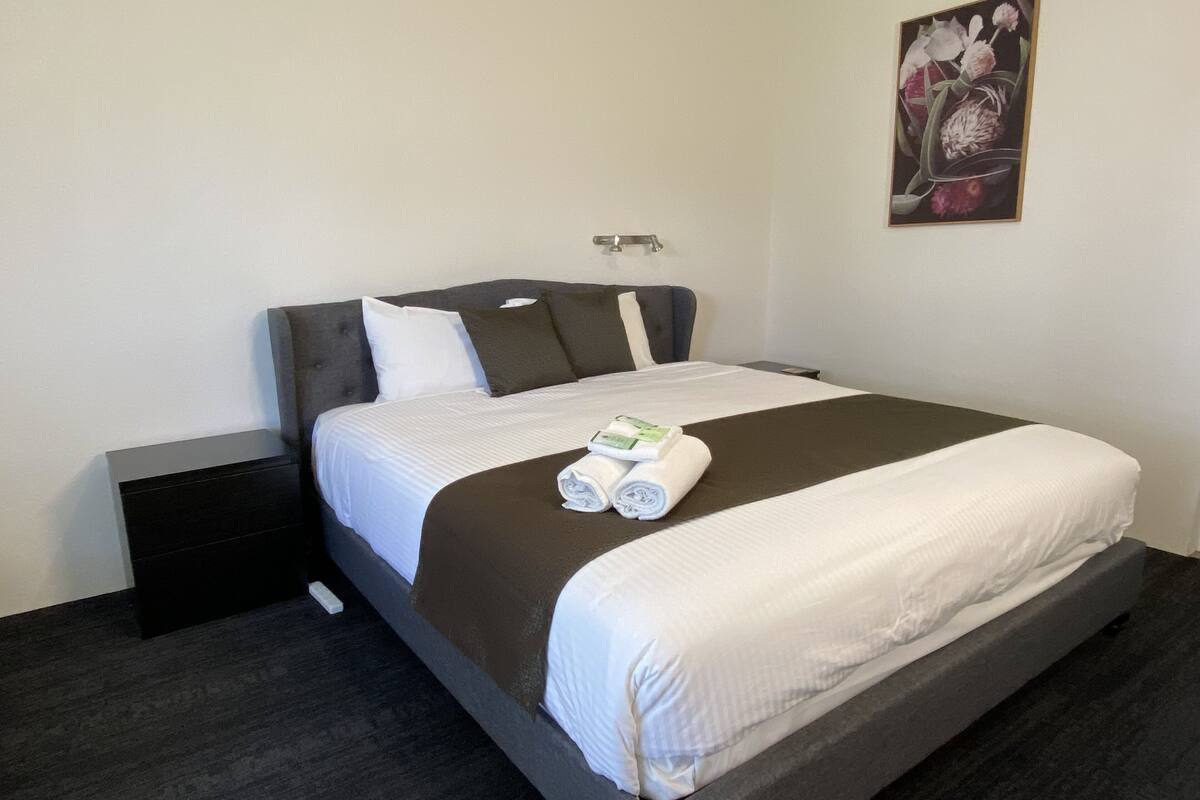 Cama bem arrumada com lençóis brancos e castanhos, uma pequena mesa de cabeceira e um quadro na parede.