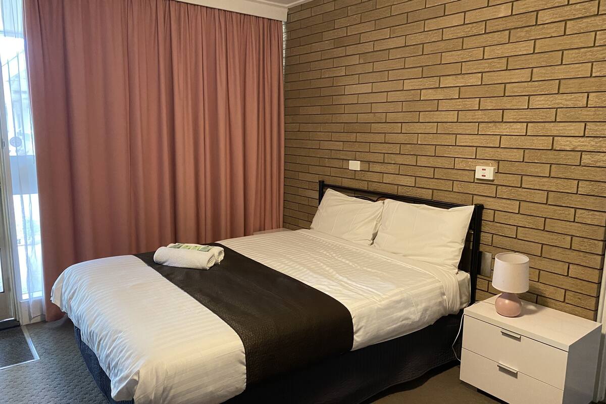Quarto de hotel com cama, mesa de cabeceira, abajur e parede de tijolos.
