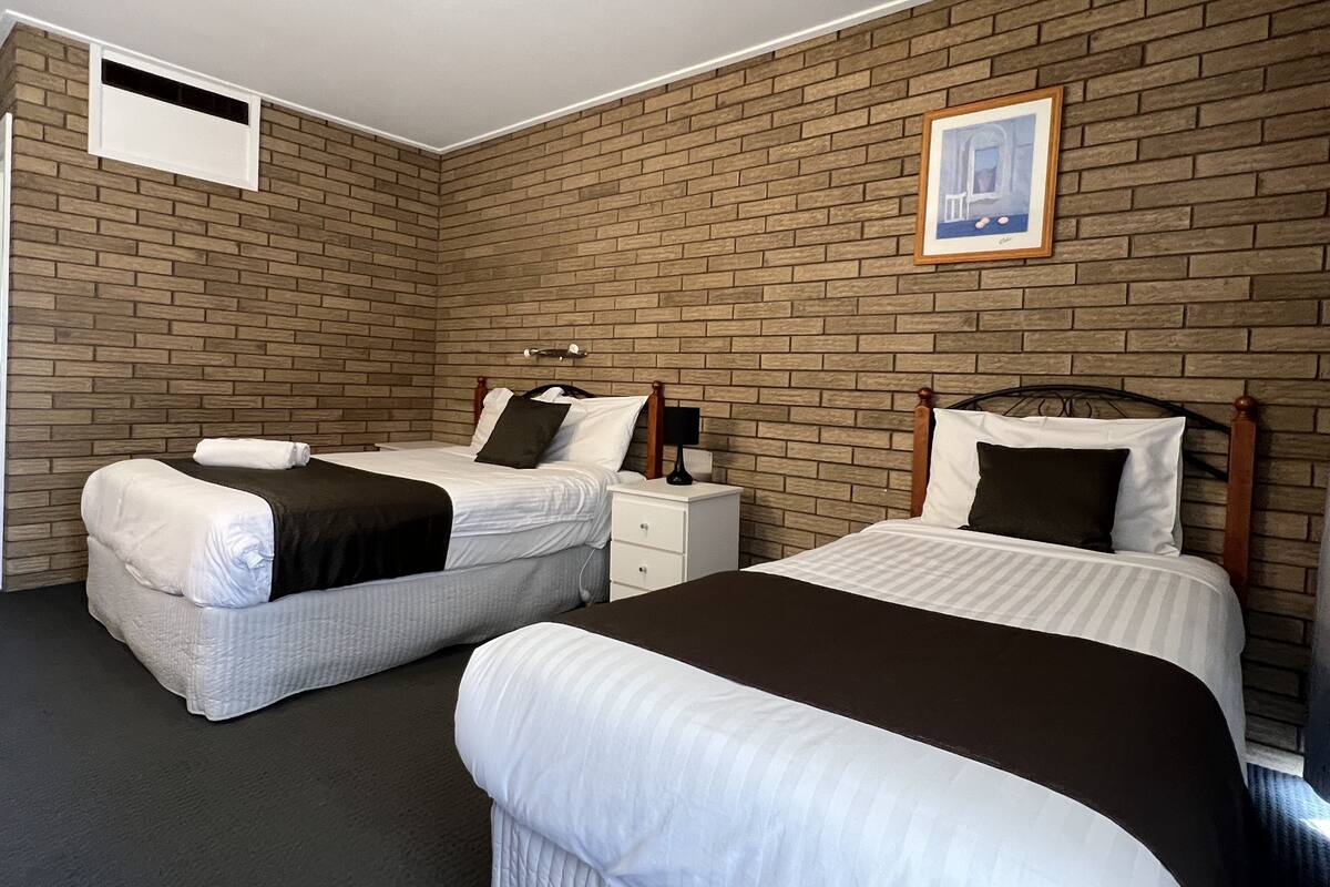 Quarto com duas camas, um penteou branco, uma mesinha e parede com padrão de tijolos.