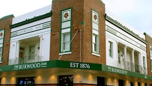 O Burwood Inn, um edifício de dois andares com fachada de tijolos e detalhes em verde. O nome do estabelecimento está em destaque na frente, e é indicado que foi fundado em 1876.