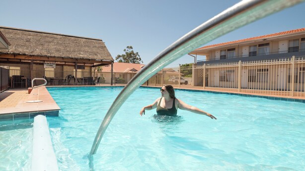 Piscina com uma pessoa na água, um edifício com telhado de palha e uma área cercada com um prédio ao fundo.