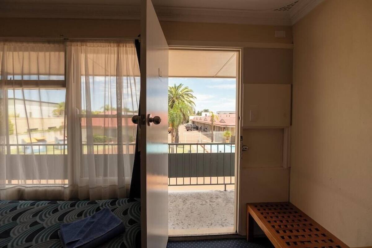 Quarto com cama, um banco, porta que dá para área externa com vista para palmeiras e edifícios.
