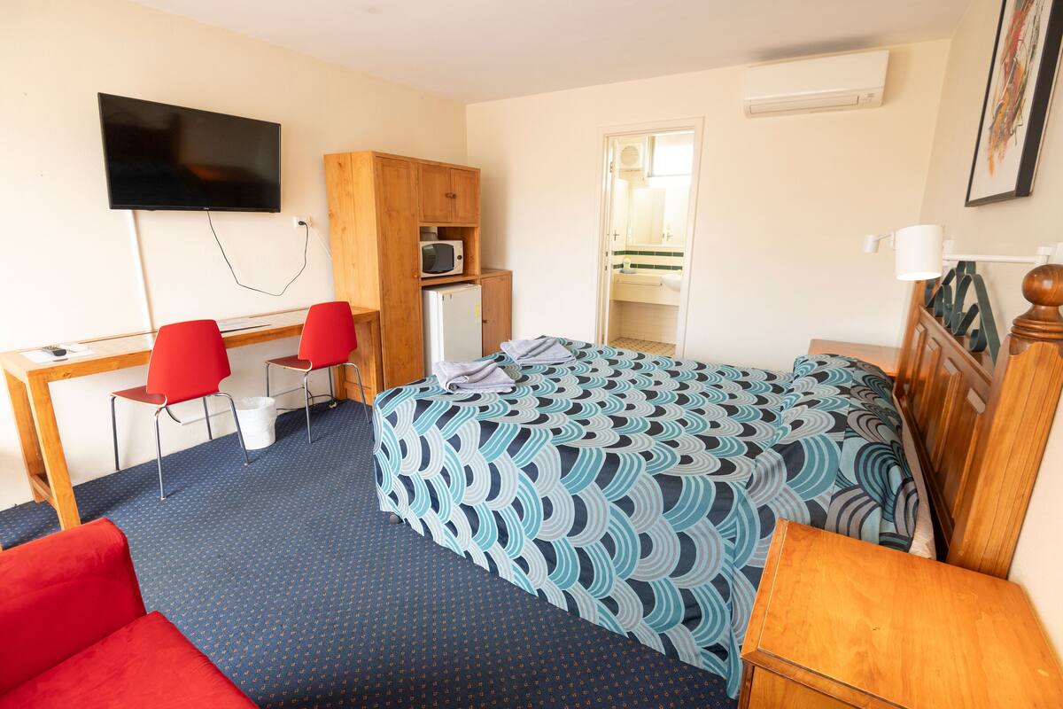 Quarto de hotel com cama, escrivaninha com cadeira, televisão, micro-ondas e um pequeno armário.