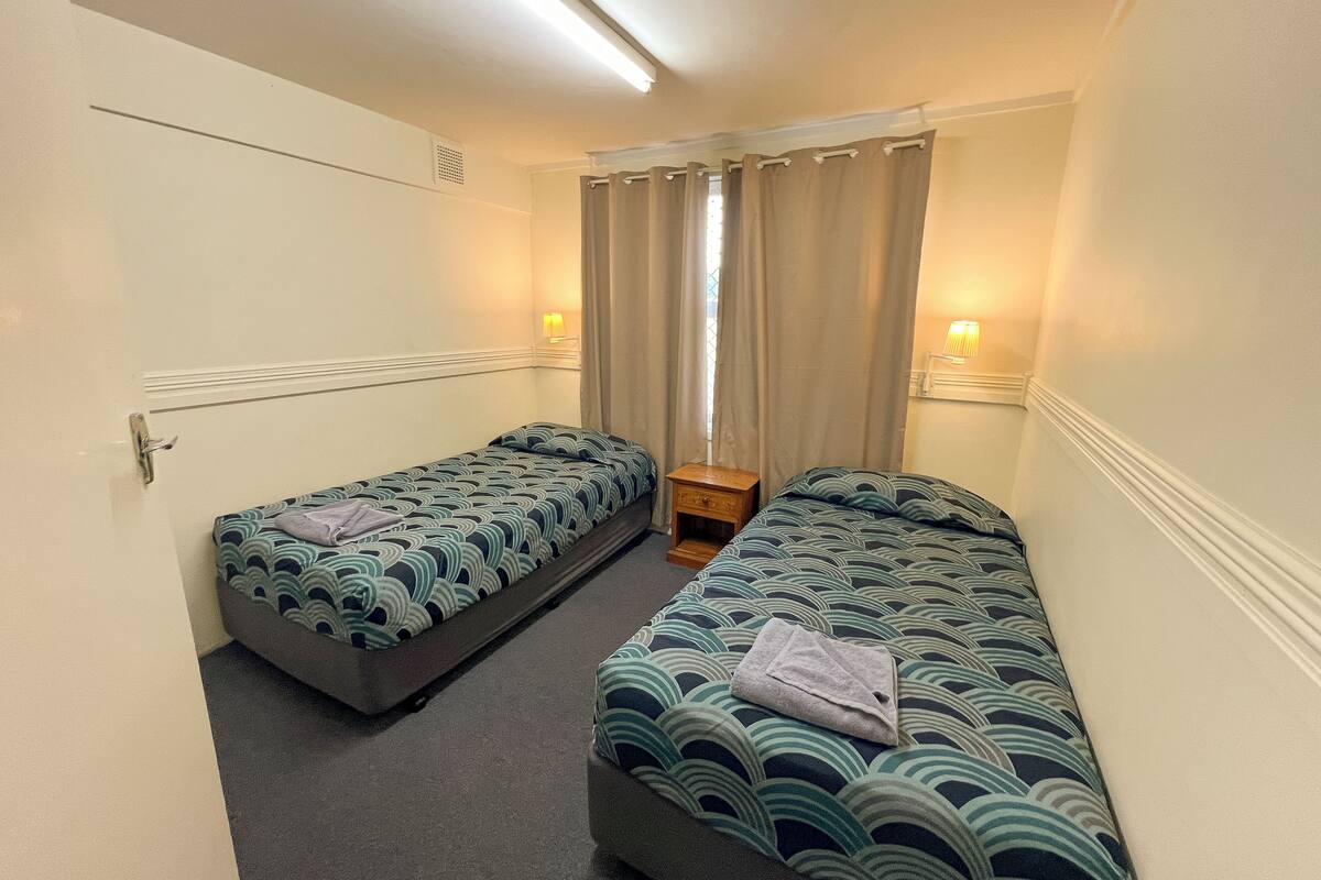 Quarto pequeno e bem iluminado com duas camas de solteiro, uma pequena mesa de cabeceira de madeira e uma janela com cortinas.