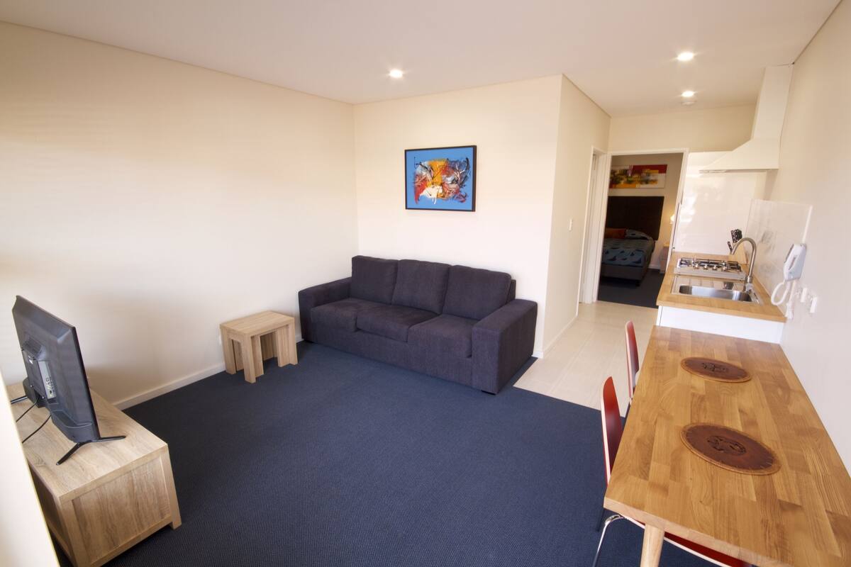 Uma sala de estar moderna com carpete azul escuro, sofá cinza, mesa de centro de madeira, televisão em suporte de madeira e um quadro na parede.