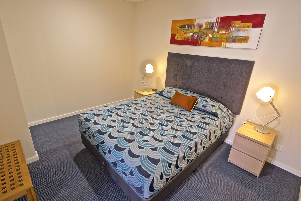 Um quarto bem arrumado com colcha estampada, cabeceira, duas mesas de cabeceira com abajures e um banquinho de madeira.