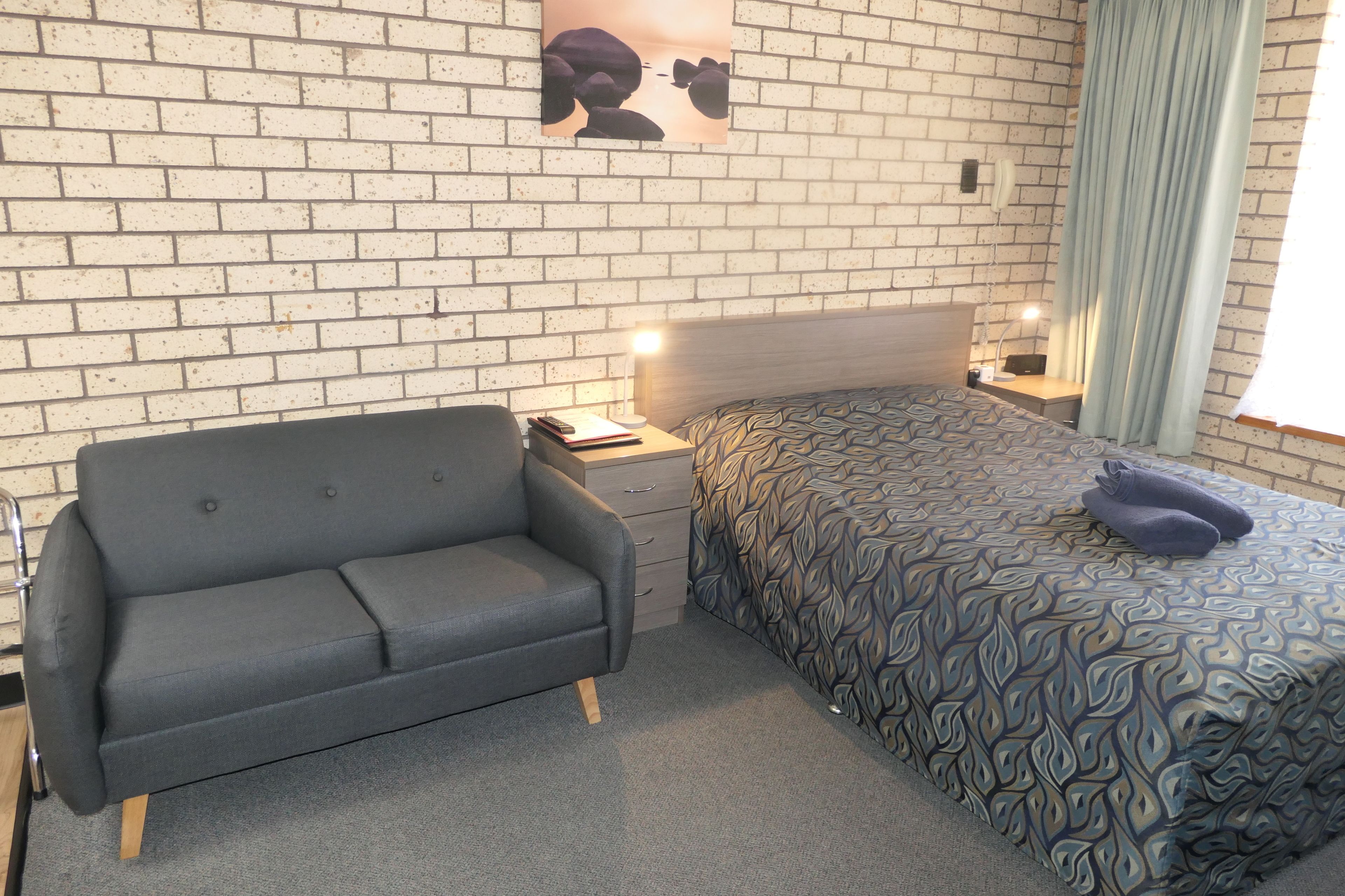 Quarto com parede de tijolos, sofá cinza, cama estampada, mesa de cabeceira com abajur e janela com cortinas.