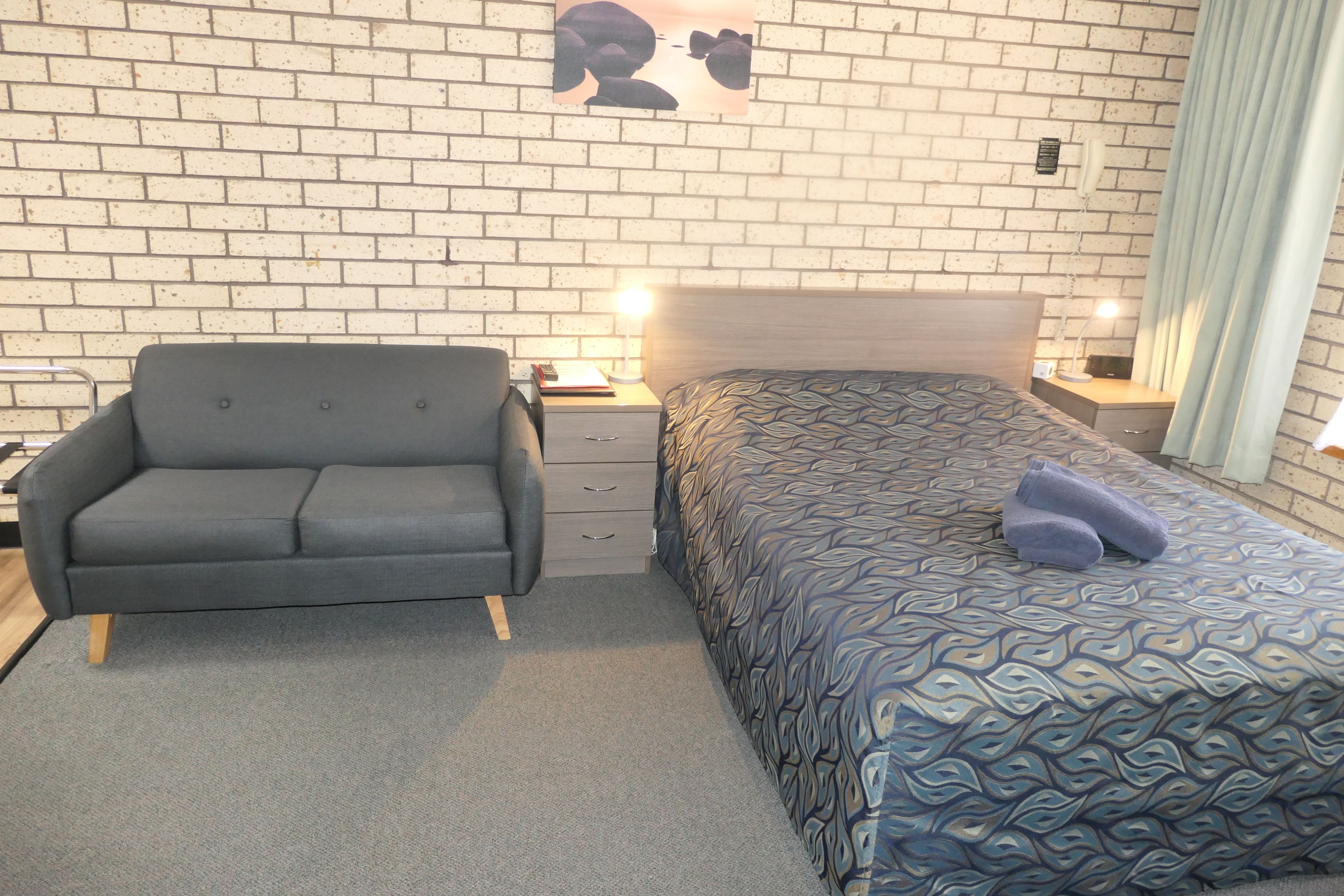 Quarto com parede de tijolos aparentes, sofá cinza, cama estampada e mesa de cabeceira com abajur.