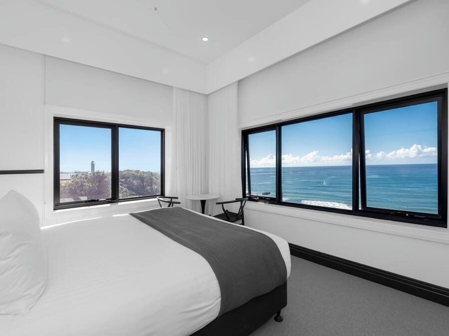 Quarto de hotel moderno com uma cama grande, duas janelas vista para o mar, uma mesinha e uma cadeira.
