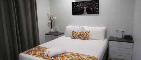 Quarto de hotel com cama, mesa de cabeceira, pequena mesa, ar condicionado e um quadro na parede.