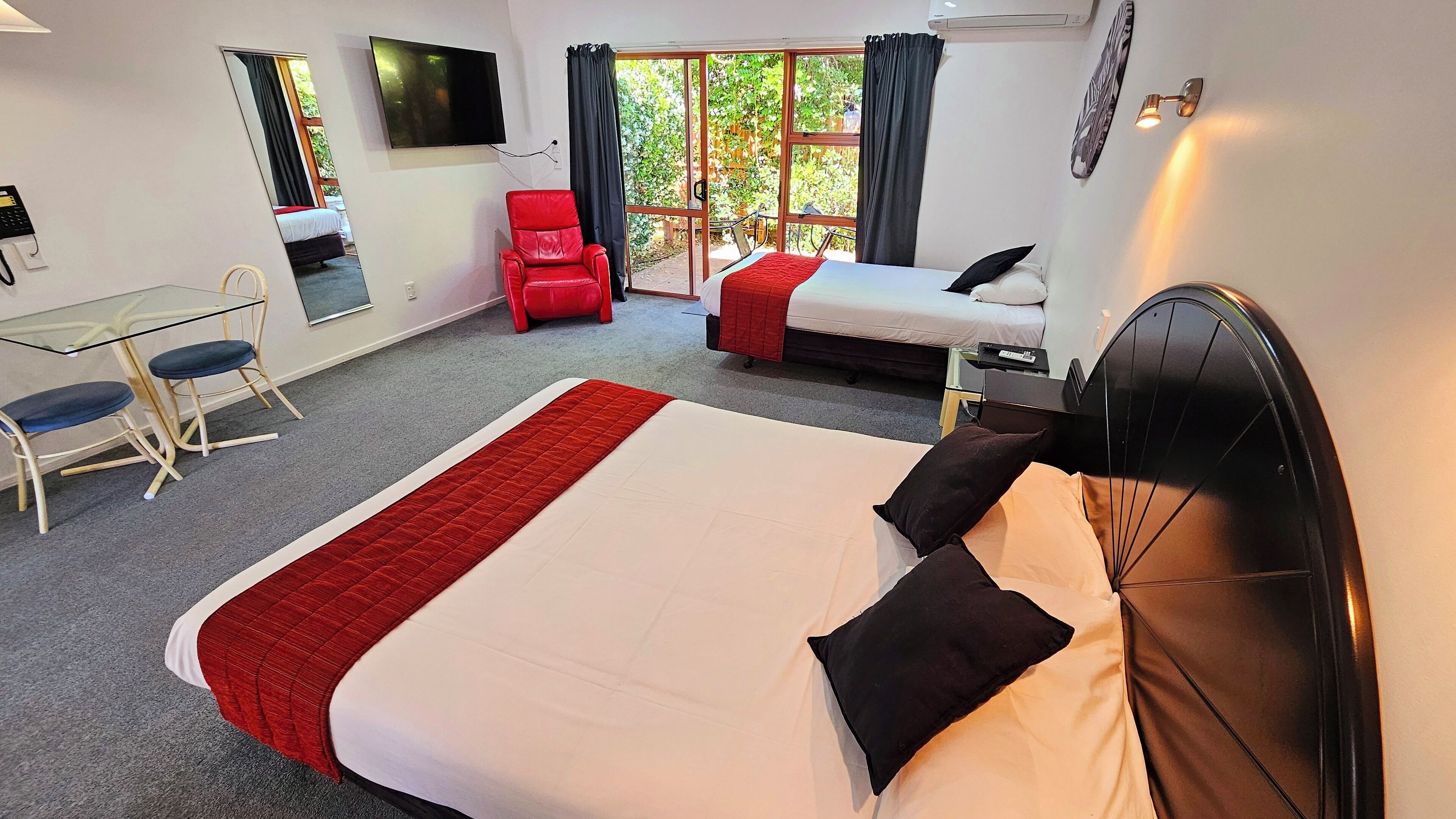 Quarto de hotel com uma cama grande, uma escrivaninha com cadeira, uma cadeira vermelha, uma televisão e uma janela com cortinas.