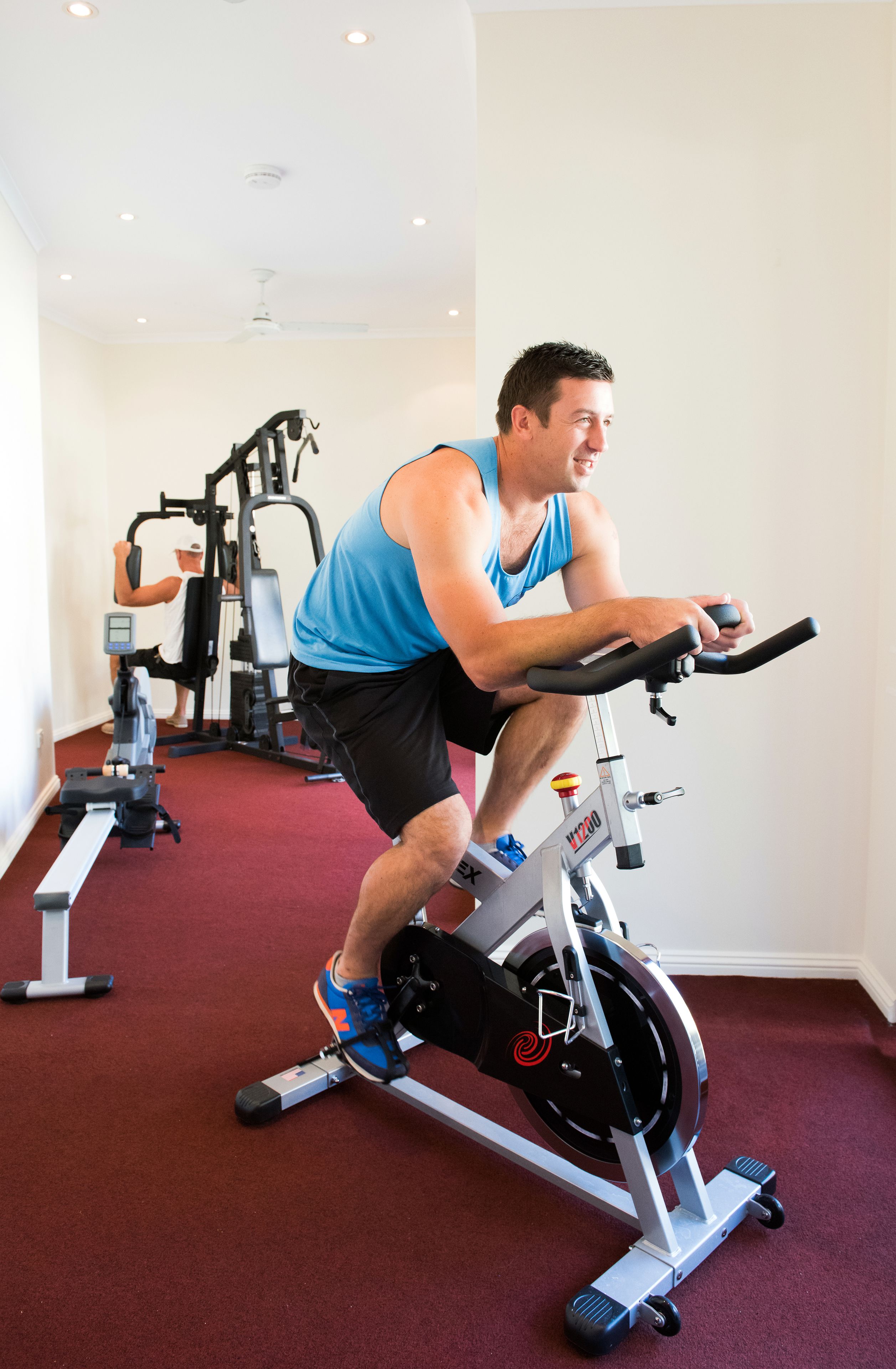 Uma pessoa pedala numa bicicleta ergométrica numa academia com piso de carpete vermelho e um ventilador de teto.