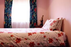 Quarto com colcha florida, uma estátua em uma mesa de cabeceira e cortinas estampadas com flores.