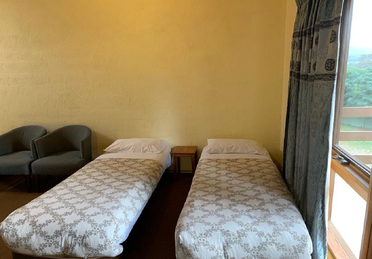 Quarto com duas camas de solteiro, uma mesinha, duas cadeiras e uma janela com cortinas.