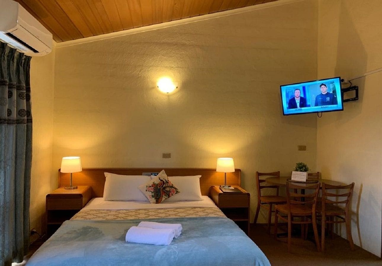 Quarto de hotel com cama, duas luminárias de cabeceira, televisão fixada na parede e uma pequena mesa com cadeiras.