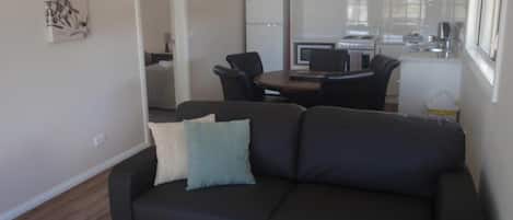 Uma sala moderna com um sofá escuro, uma mesa de jantar com cadeiras e uma área de cozinha com armários e eletrodomésticos brancos.