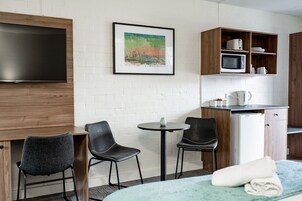 Quarto de hotel com uma cama, duas cadeiras, uma mesa pequena, televisão, micro-ondas e um quadro na parede.