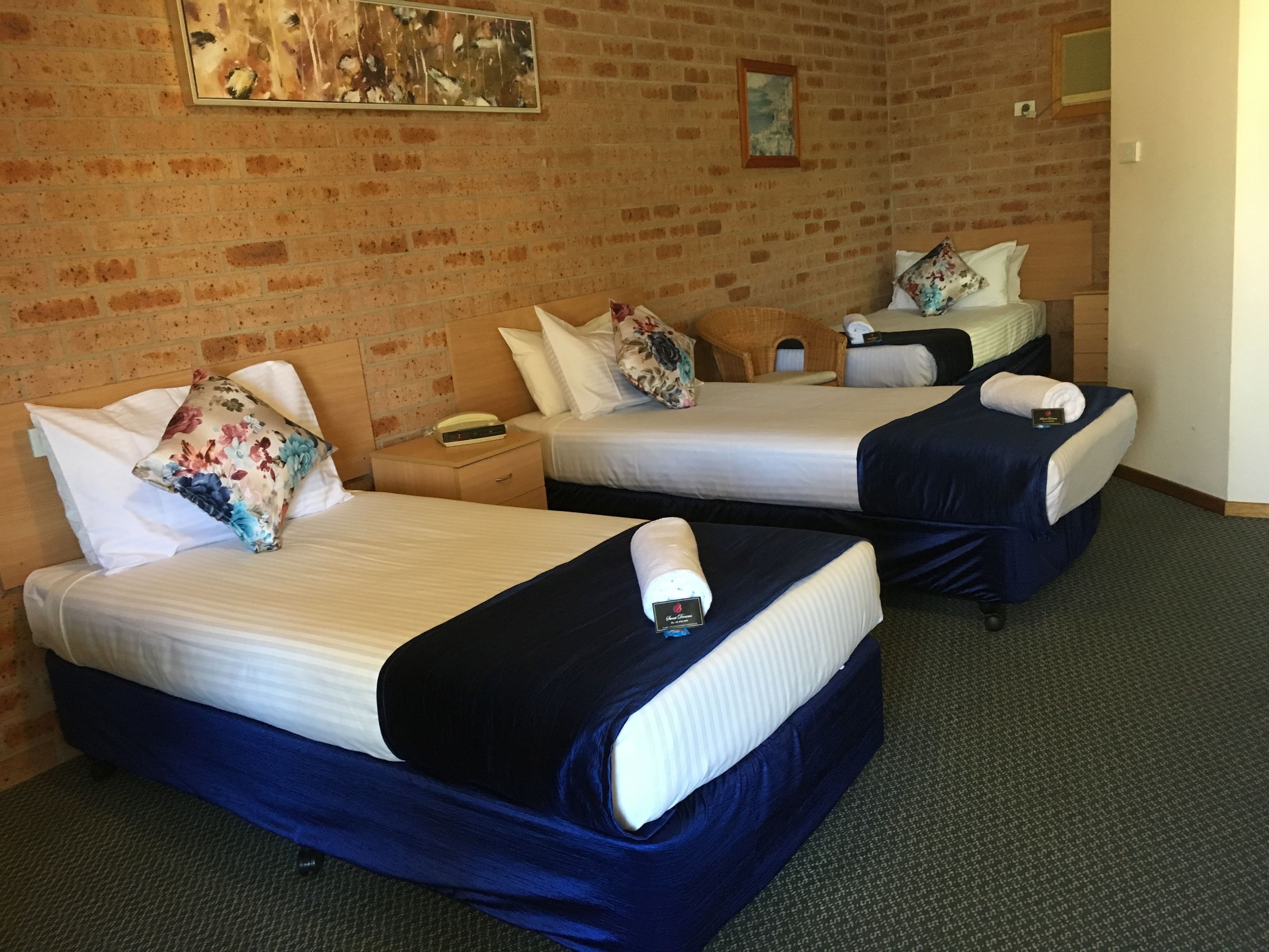 Quarto de hotel com duas camas, cada uma com travesseiro e cobertor. Há uma mesinha de cabeceira com um telefone e uma pequena mesa entre as camas. O quarto tem paredes de tijolo e um quadro na parede.