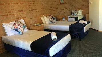 Quarto de hotel com duas camas, cada uma com travesseiro e cobertor. Há uma mesinha de cabeceira com um telefone e uma pequena mesa entre as camas. O quarto tem paredes de tijolo e um quadro na parede.
