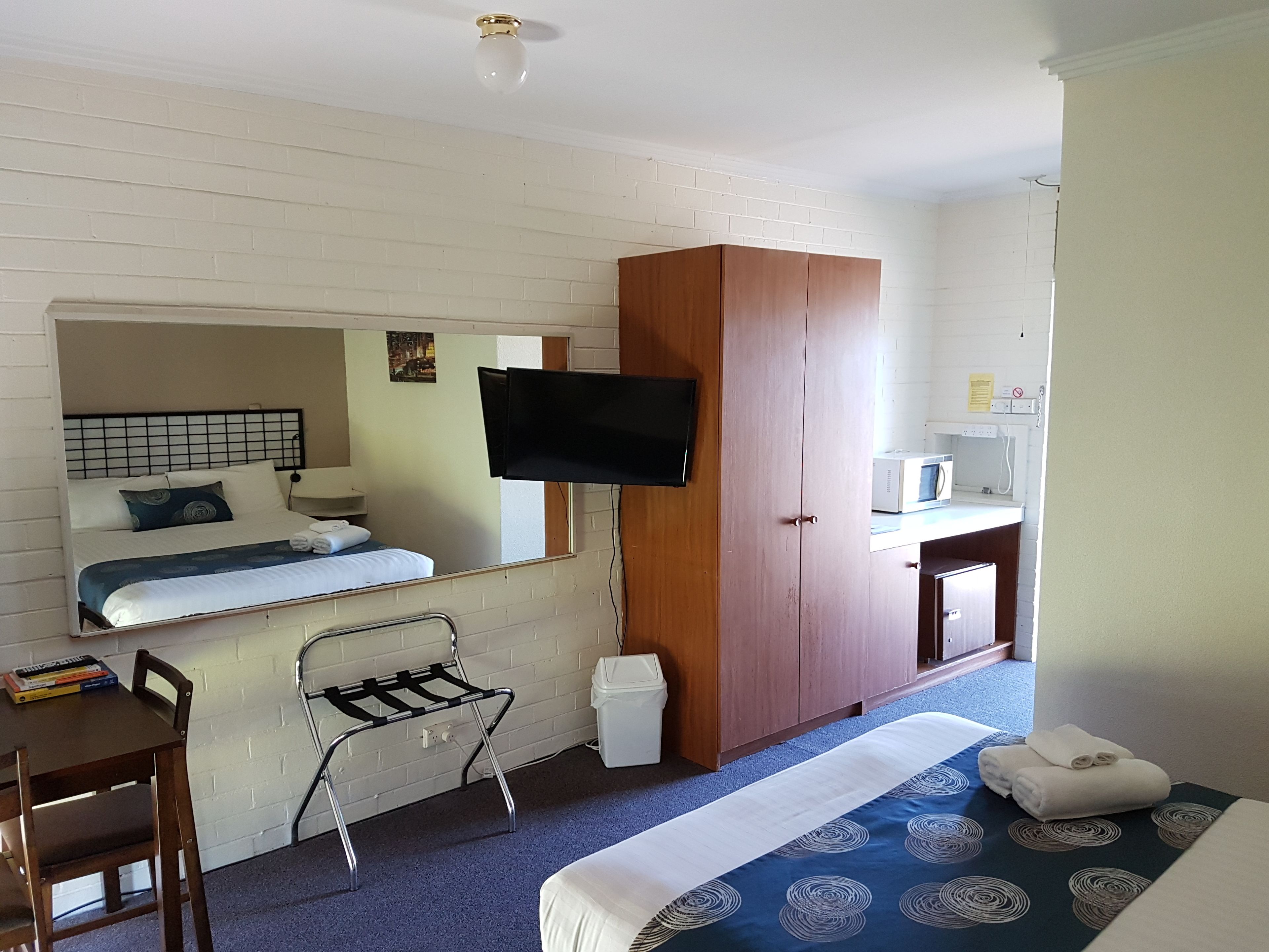Quarto de hotel com uma cama, uma mesa com computador, televisão na parede e uma pequena cozinha equipada.