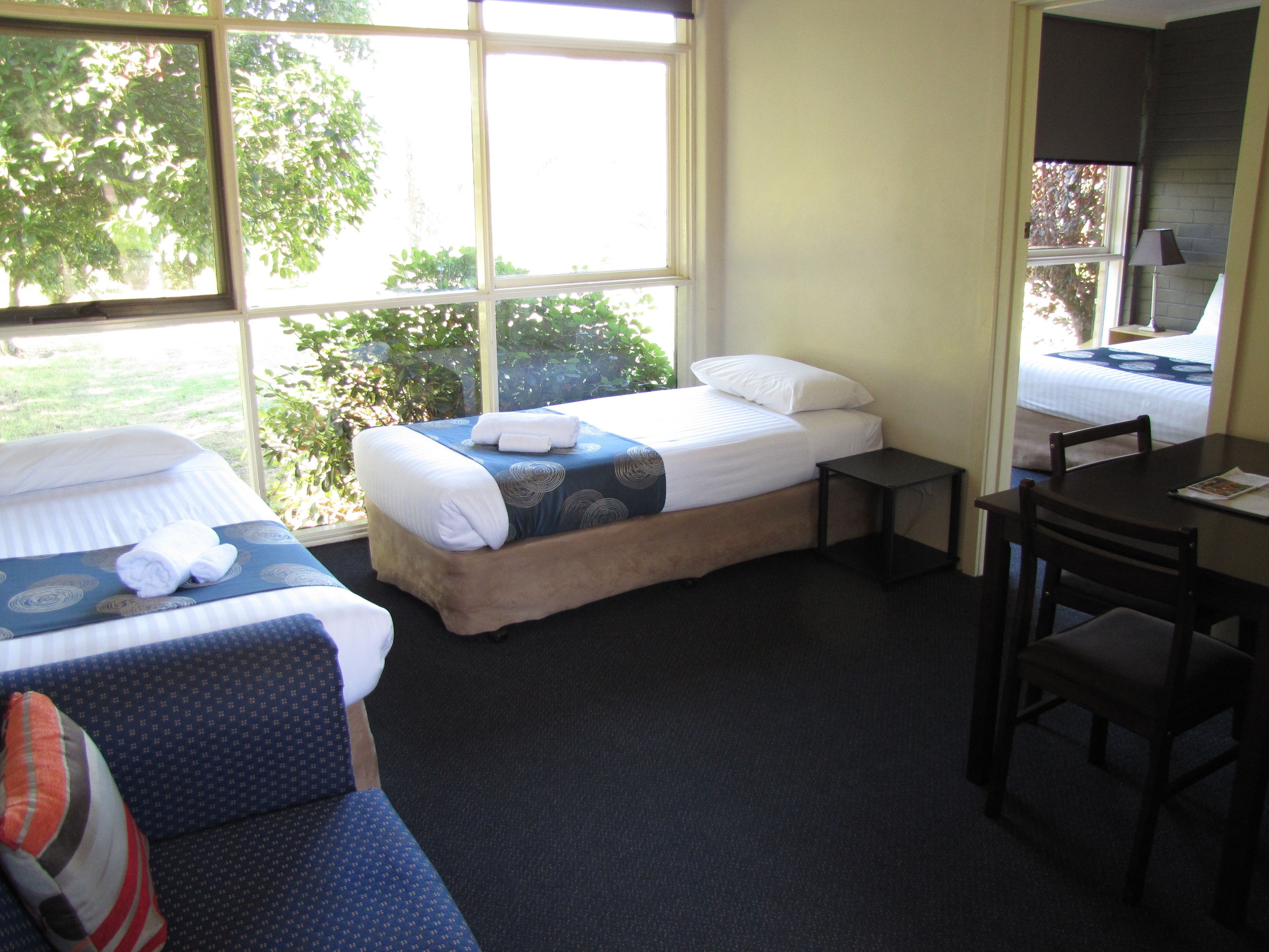 Quarto de hotel com duas camas, uma mesinha, uma cadeira e vista para o exterior.