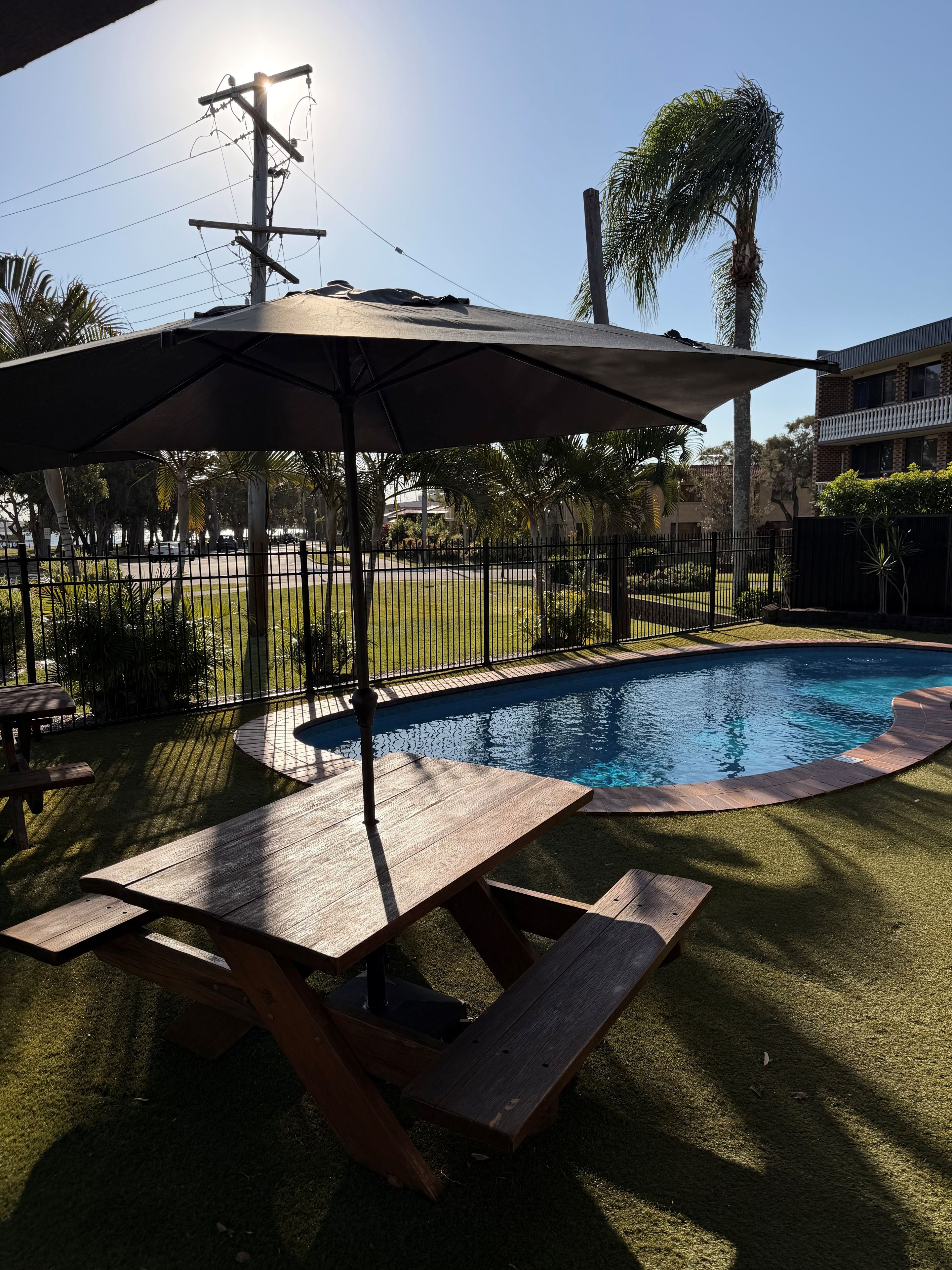 Piscina com deck de madeira e mesas de piquenique, cercada por uma grade e palmeiras.