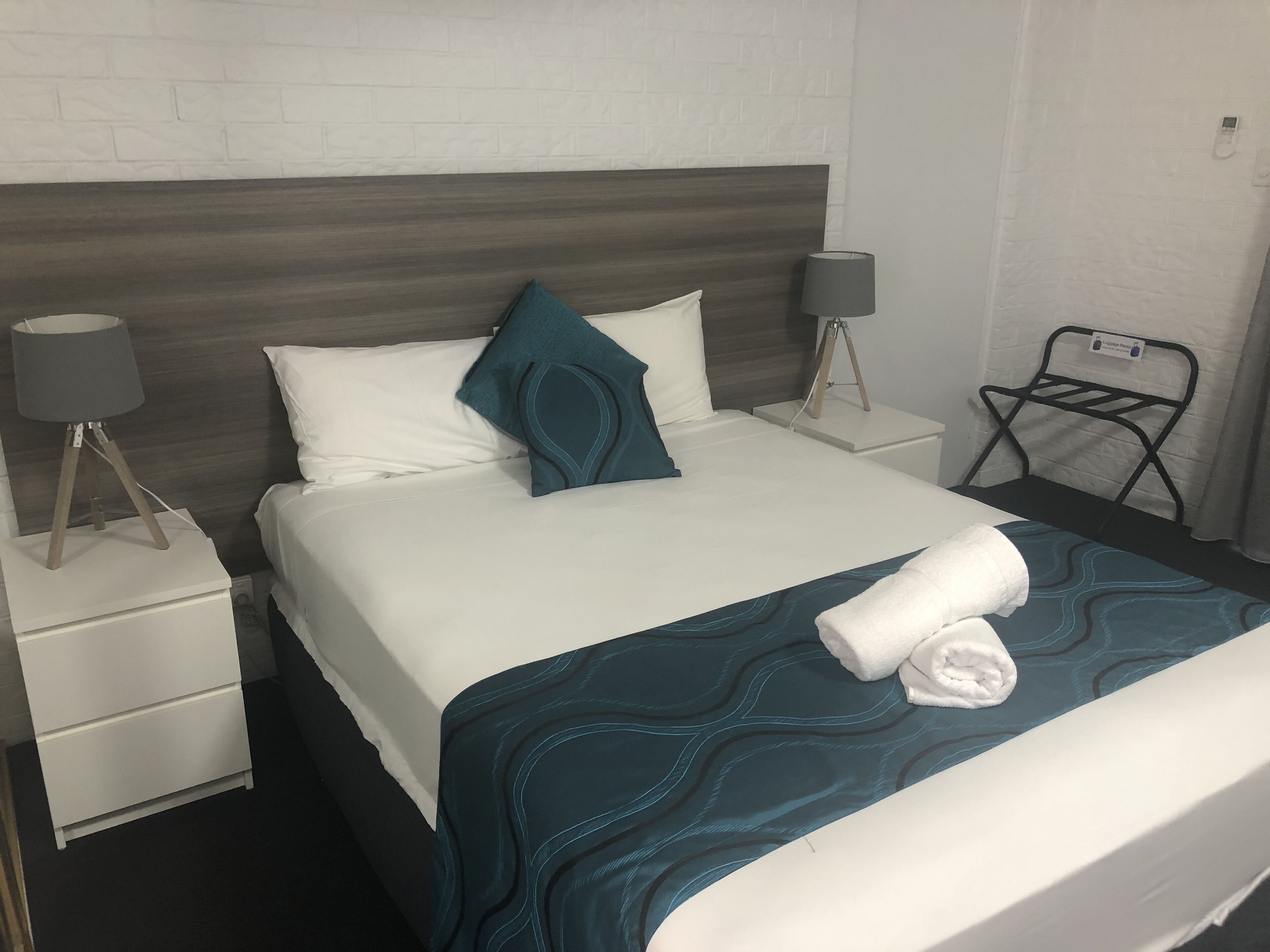 Cama bem arrumada com roupa de cama branca e azul turquesa, duas luminárias de cabeceira e uma pequena mesa de cabeceira com um abajur.