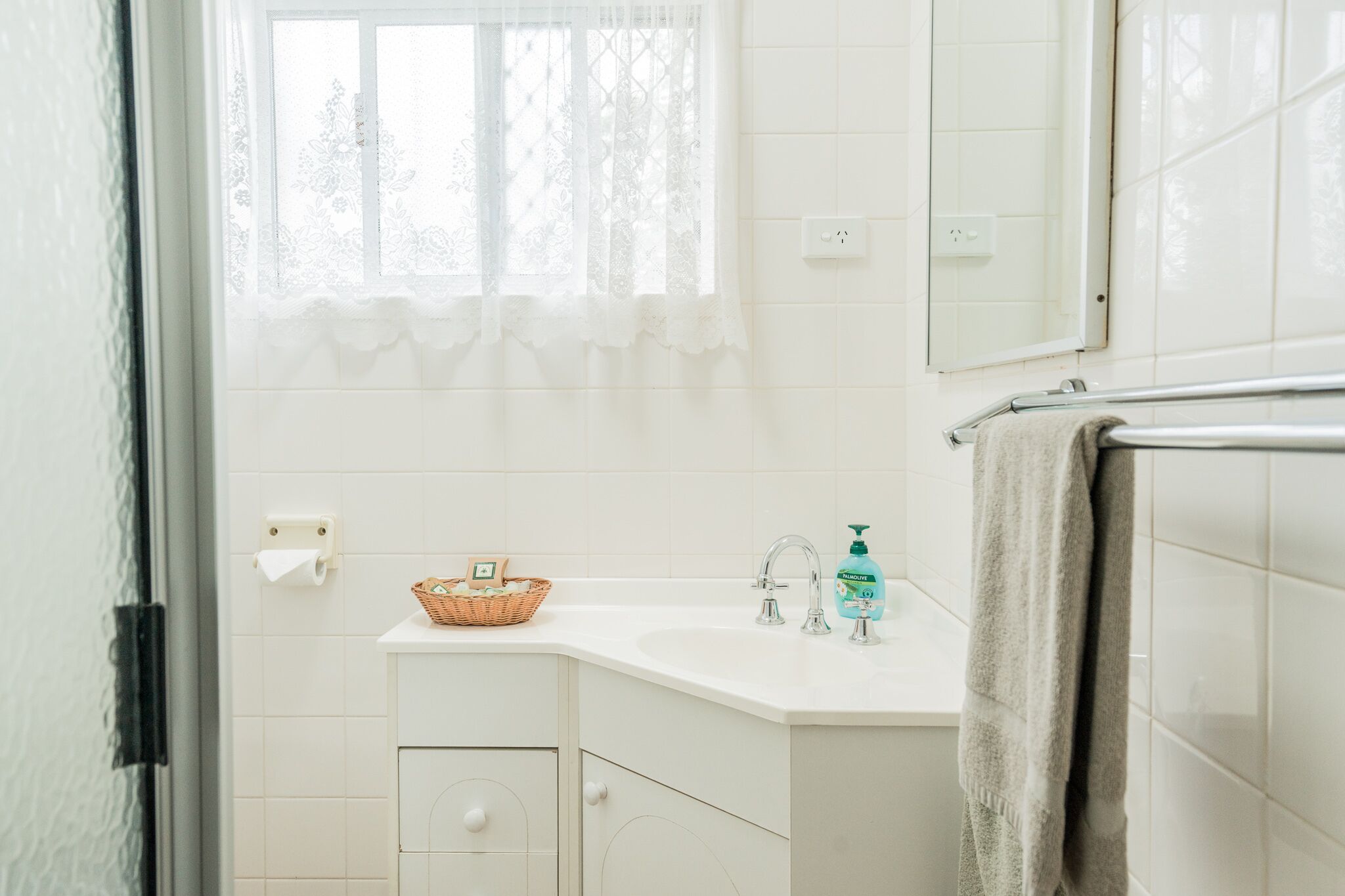 Uma casa de banho com uma pia branca, um espelho, um porta-toalhas e uma janela com cortinas de renda.