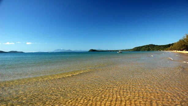 Uma praia com águas claras e rasas, e montanhas ao longe.
