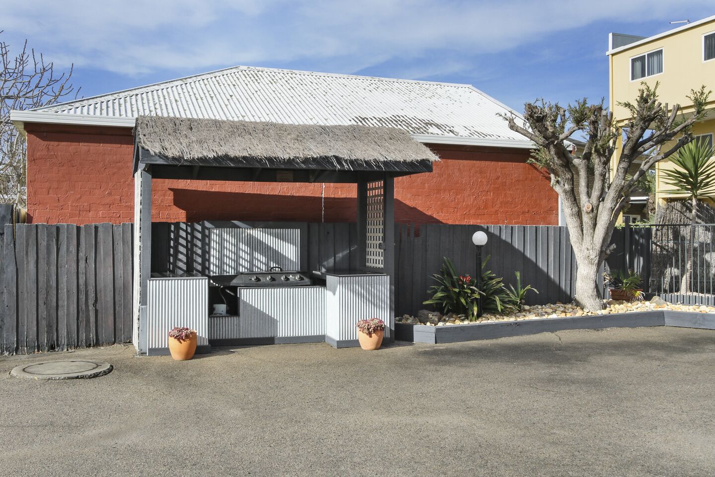 Um edifício com área de churrasco coberta ao ar livre, um quintal cercado e vasos de plantas.