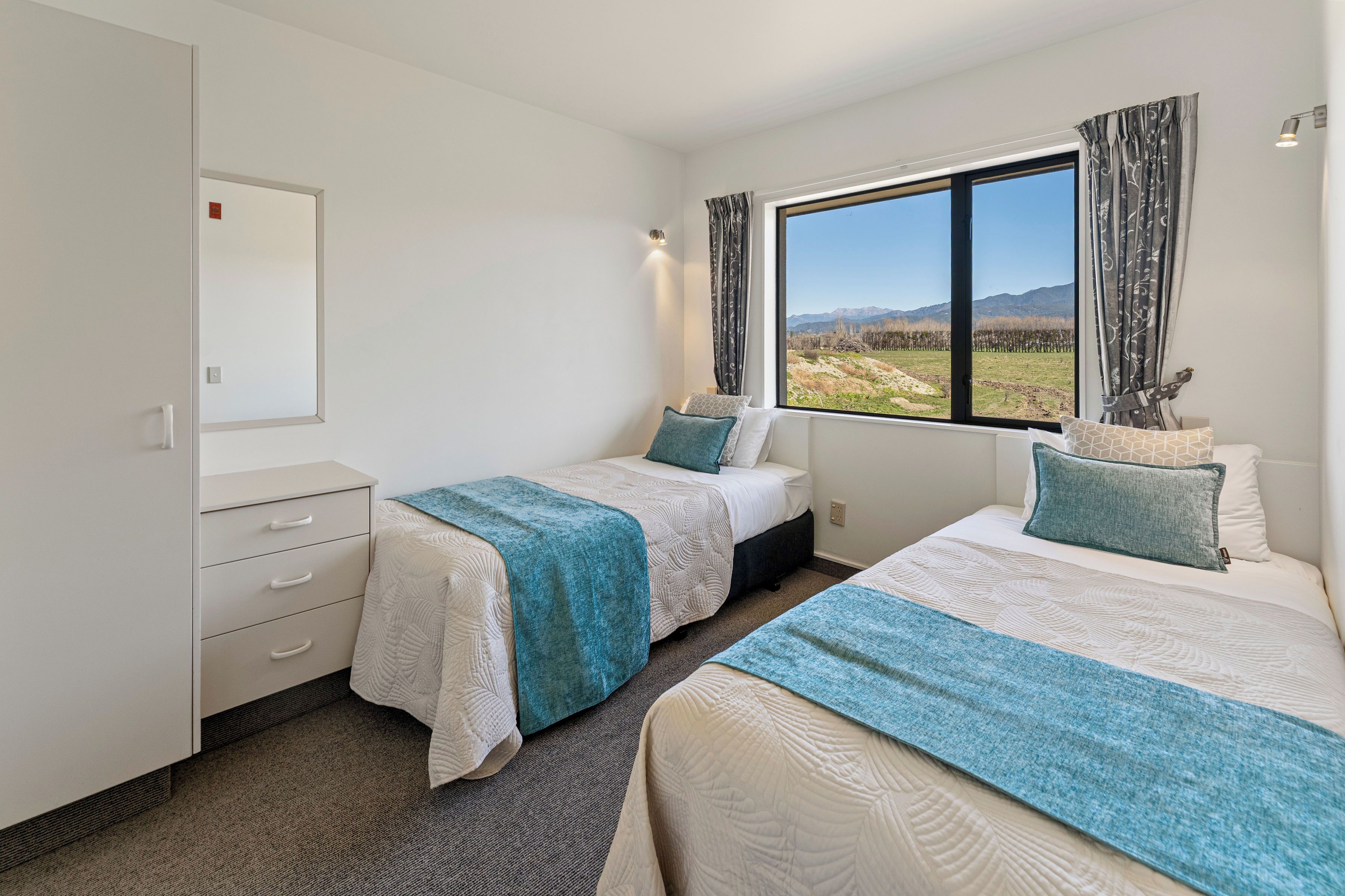 Quarto de hotel com duas camas, uma cômoda e uma janela ampla com vista para montanhas.