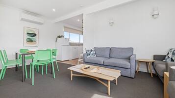 Uma sala de estar moderna com um sofá cinzento, poltronas verdes, uma mesa de centro de madeira e uma cozinha ao fundo.