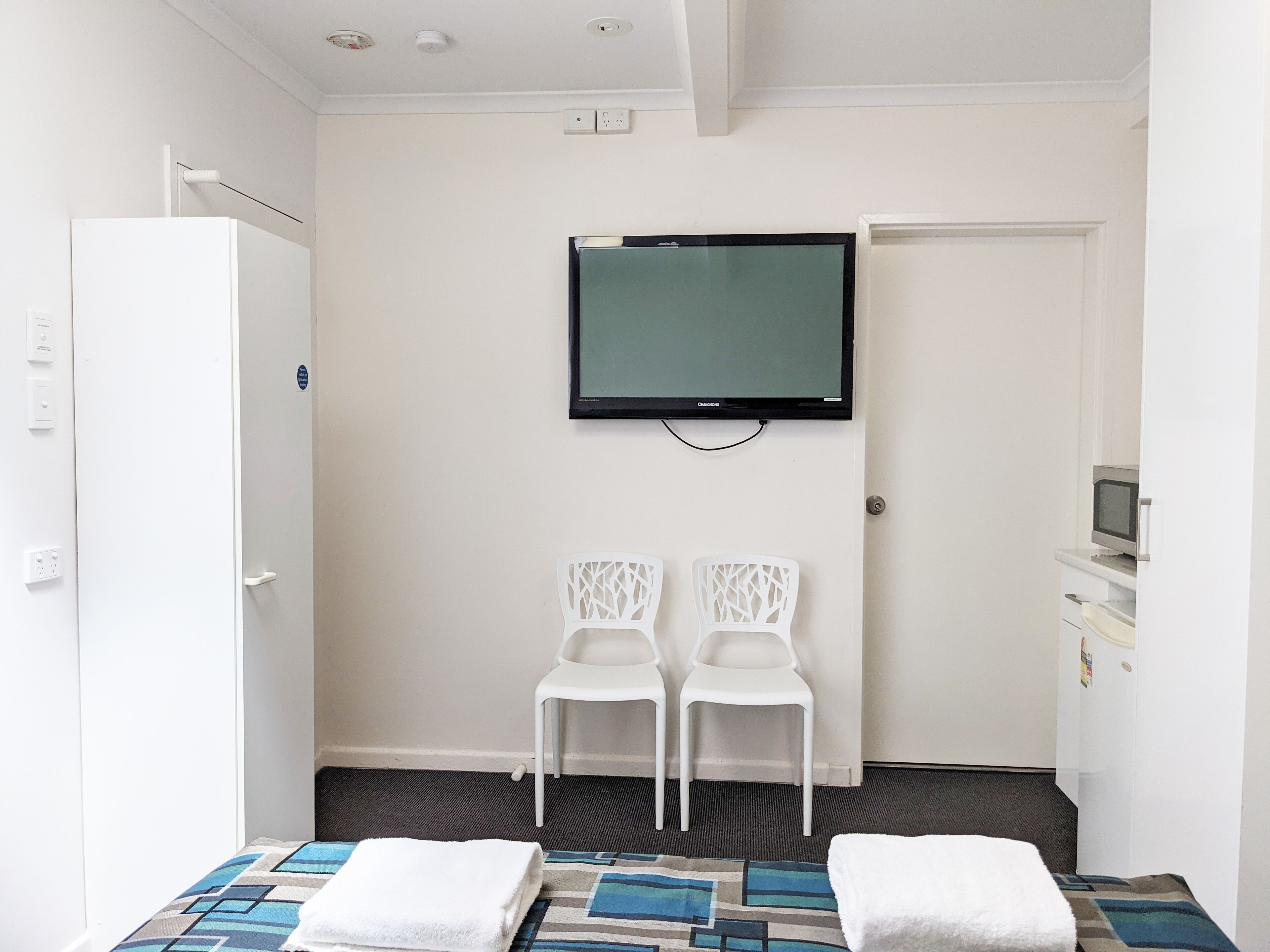 Quarto de hotel com uma cama, duas cadeiras brancas, uma televisão e um minibar.