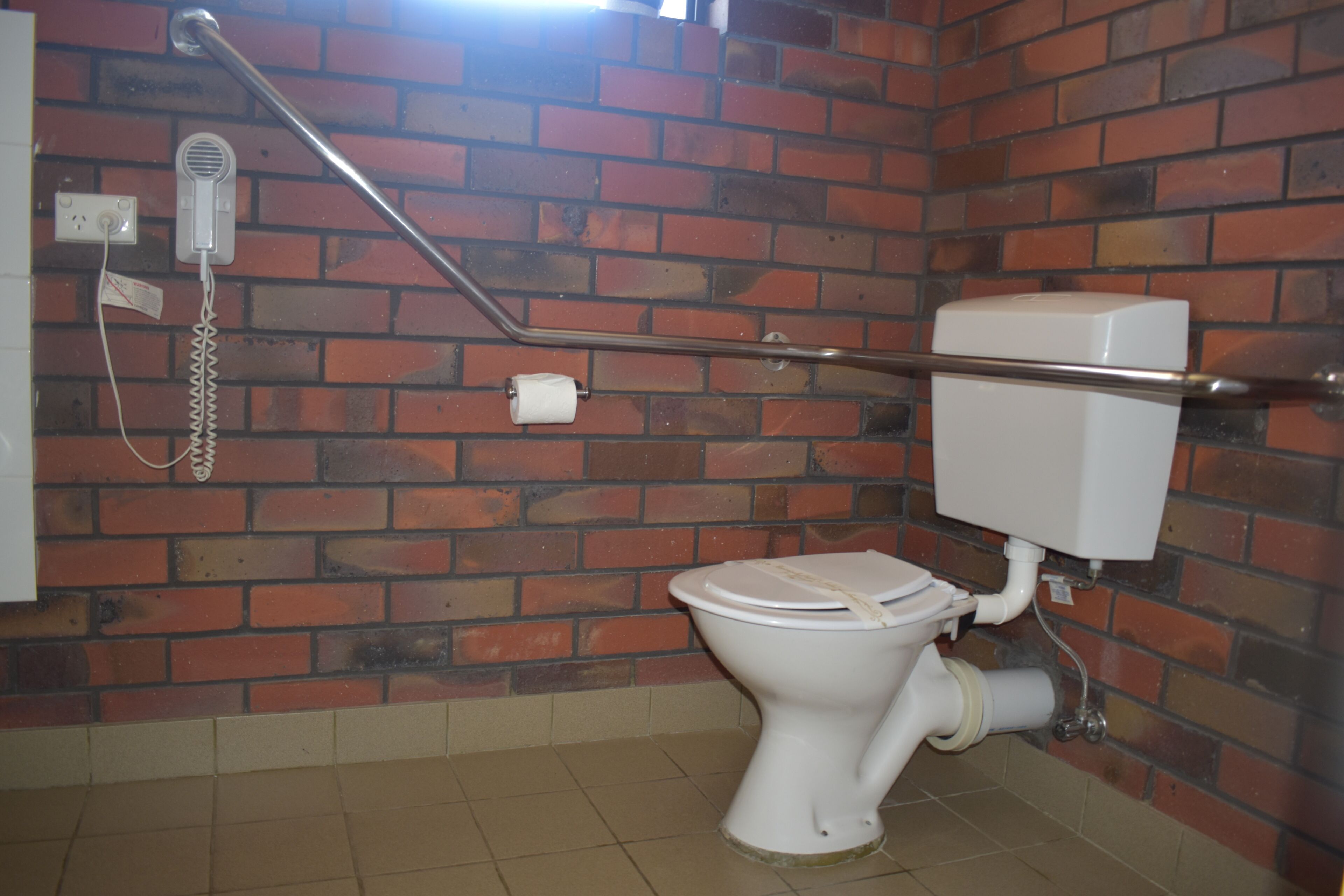 Casa de banho com sanita branca, secador de mãos e dispensador de toalhas de papel fixado numa parede de tijolo.