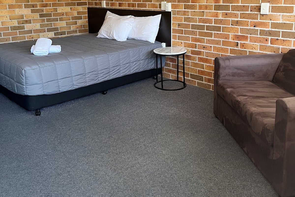 Um quarto com parede de tijolos aparentes, uma cama com colchão cinzento e almofadas brancas, uma mesinha de cabeceira redonda e um sofá marrom.