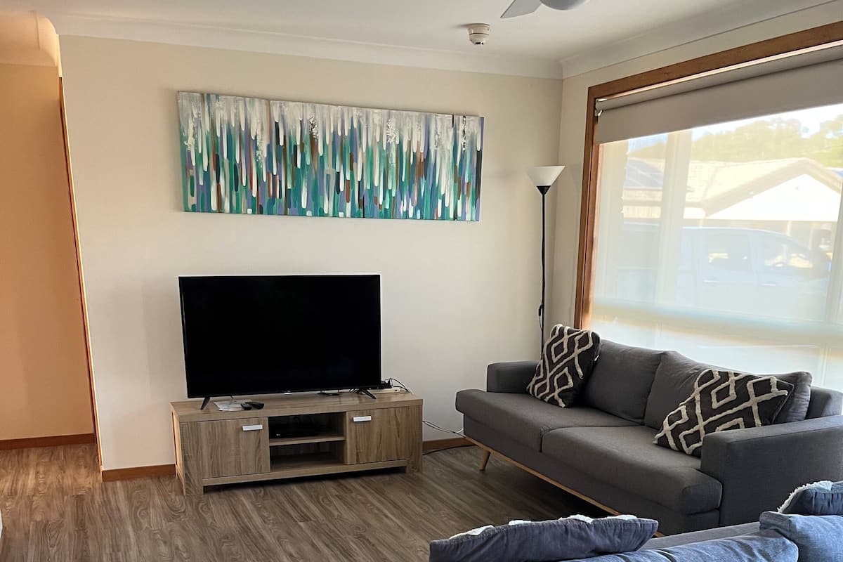 Uma sala de estar moderna com uma TV de tela plana sobre um suporte de madeira, um sofá cinza com almofadas estampadas e um grande quadro abstrato na parede.