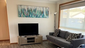Uma sala de estar moderna com uma TV de tela plana sobre um suporte de madeira, um sofá cinza com almofadas estampadas e um grande quadro abstrato na parede.